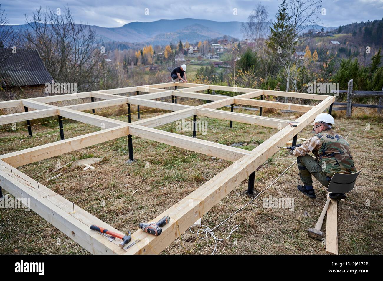 Man building wooden frame house on pile foundation. Male worker drilling hole by electric drill ...