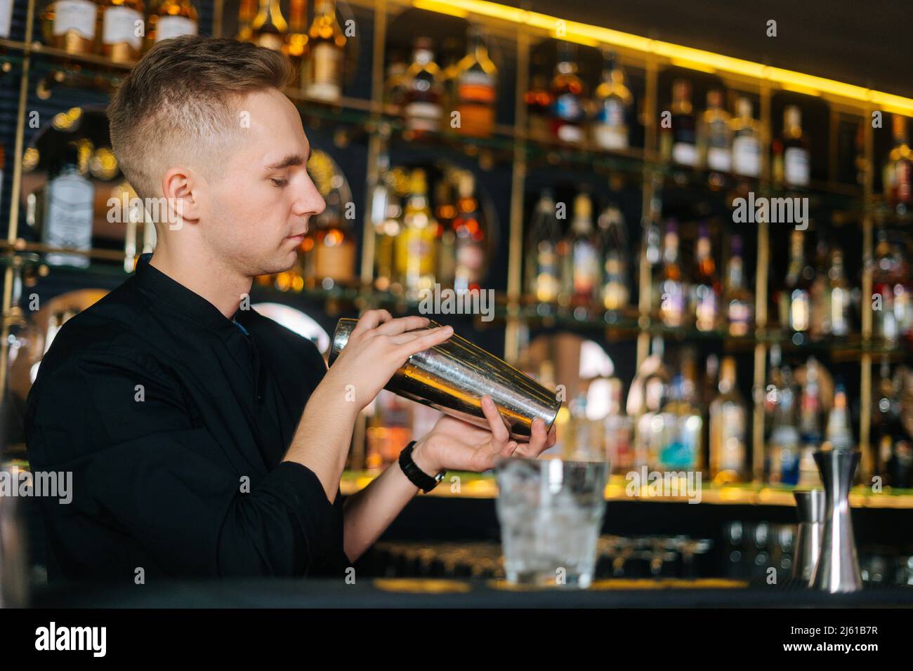 Side view of bartender mixing ingredients of alcoholic cocktail by shaking shaker standing ...