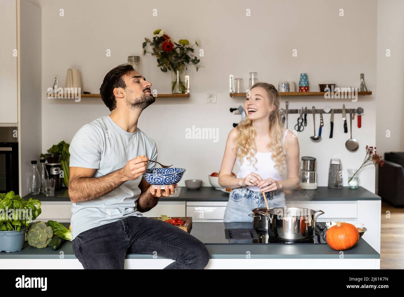 Woman looking at man eating food sitting on kitchen counter at home ...