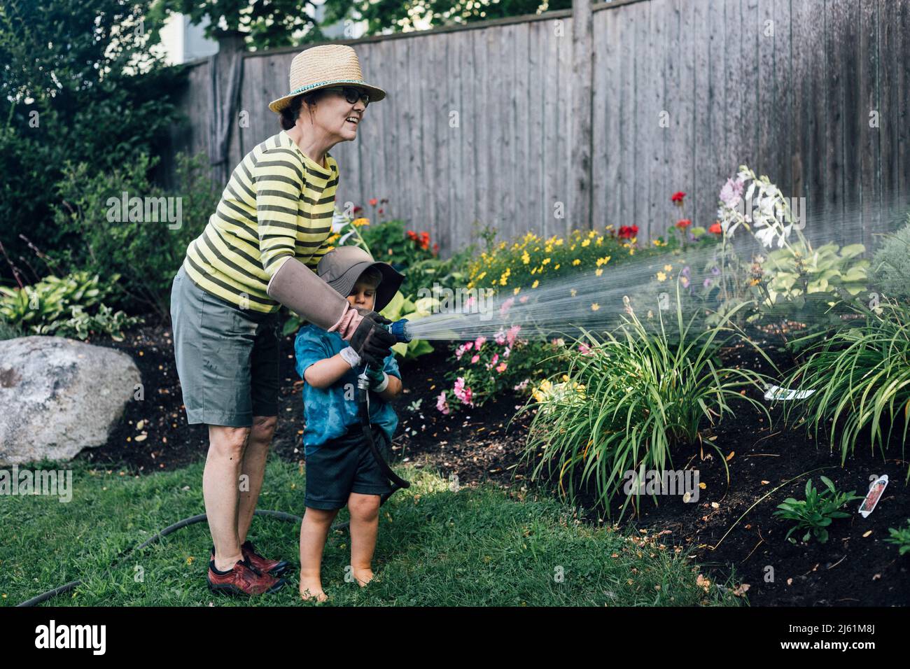 Happy Grandmother With Grandson Watering Plants Together In Garden