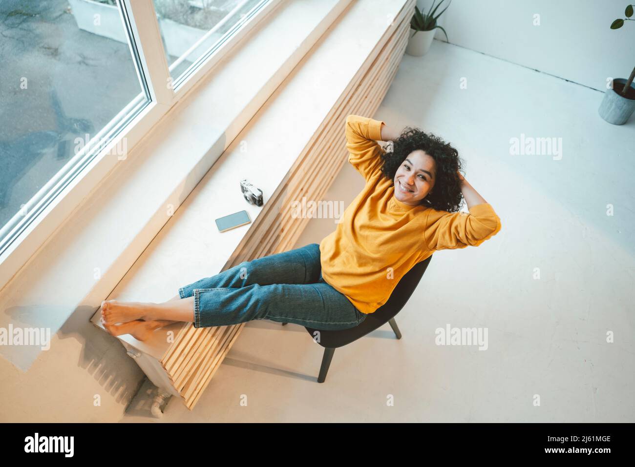 Happy woman with feet up and hands behind head sitting on chair at home ...