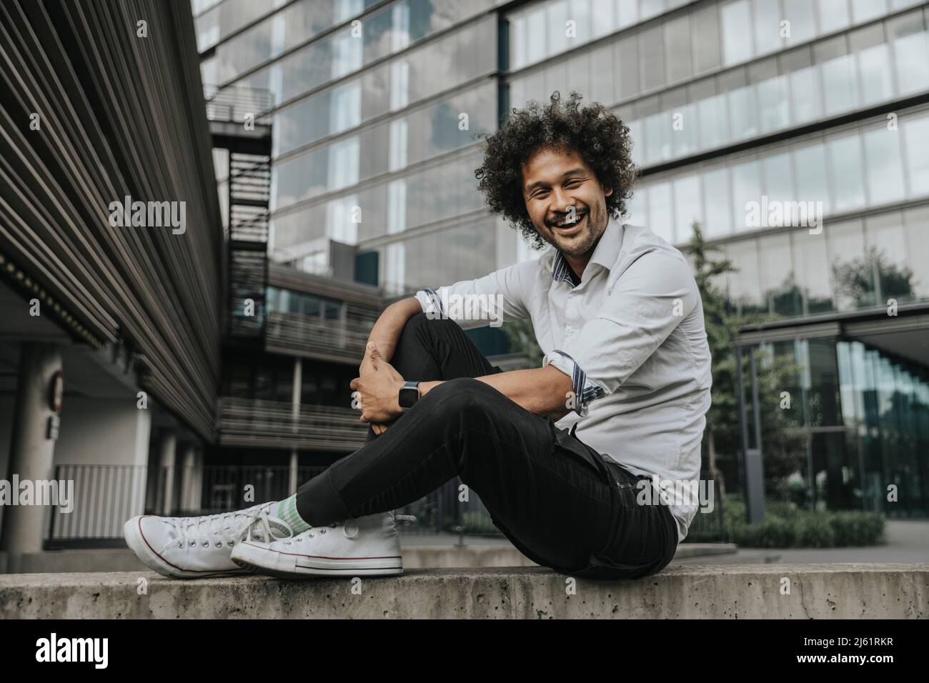 Happy young man sitting on ledge in front of modern building Stock ...