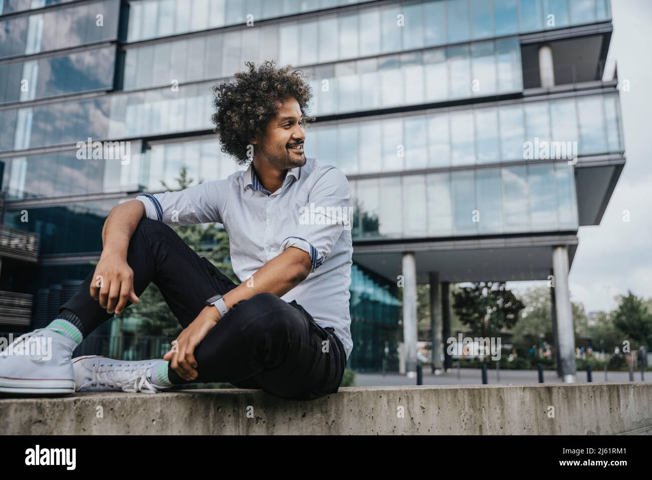 Smiling young man sitting on ledge in front of modern building Stock ...