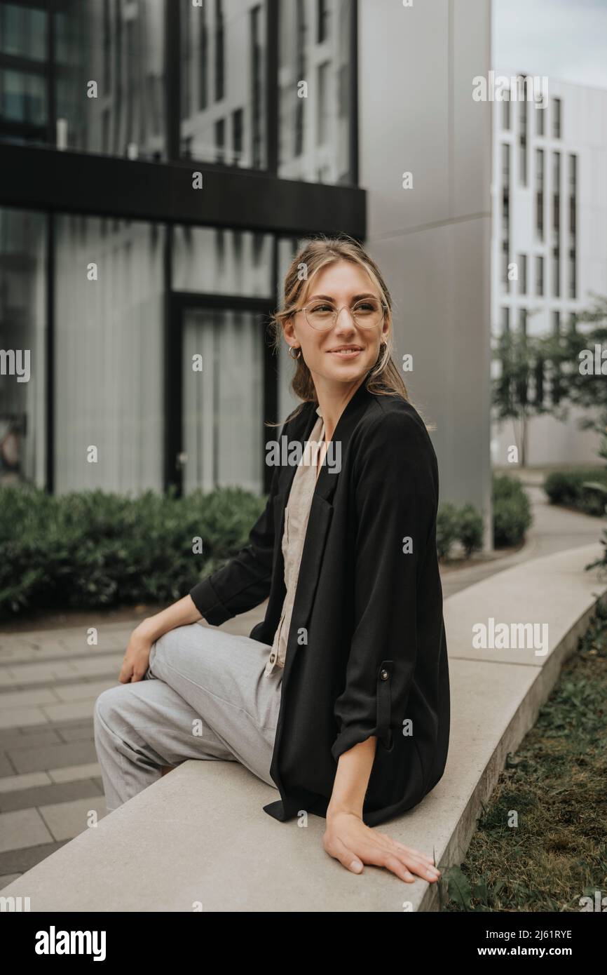 Smiling young woman sitting on ledge in front of building Stock Photo ...