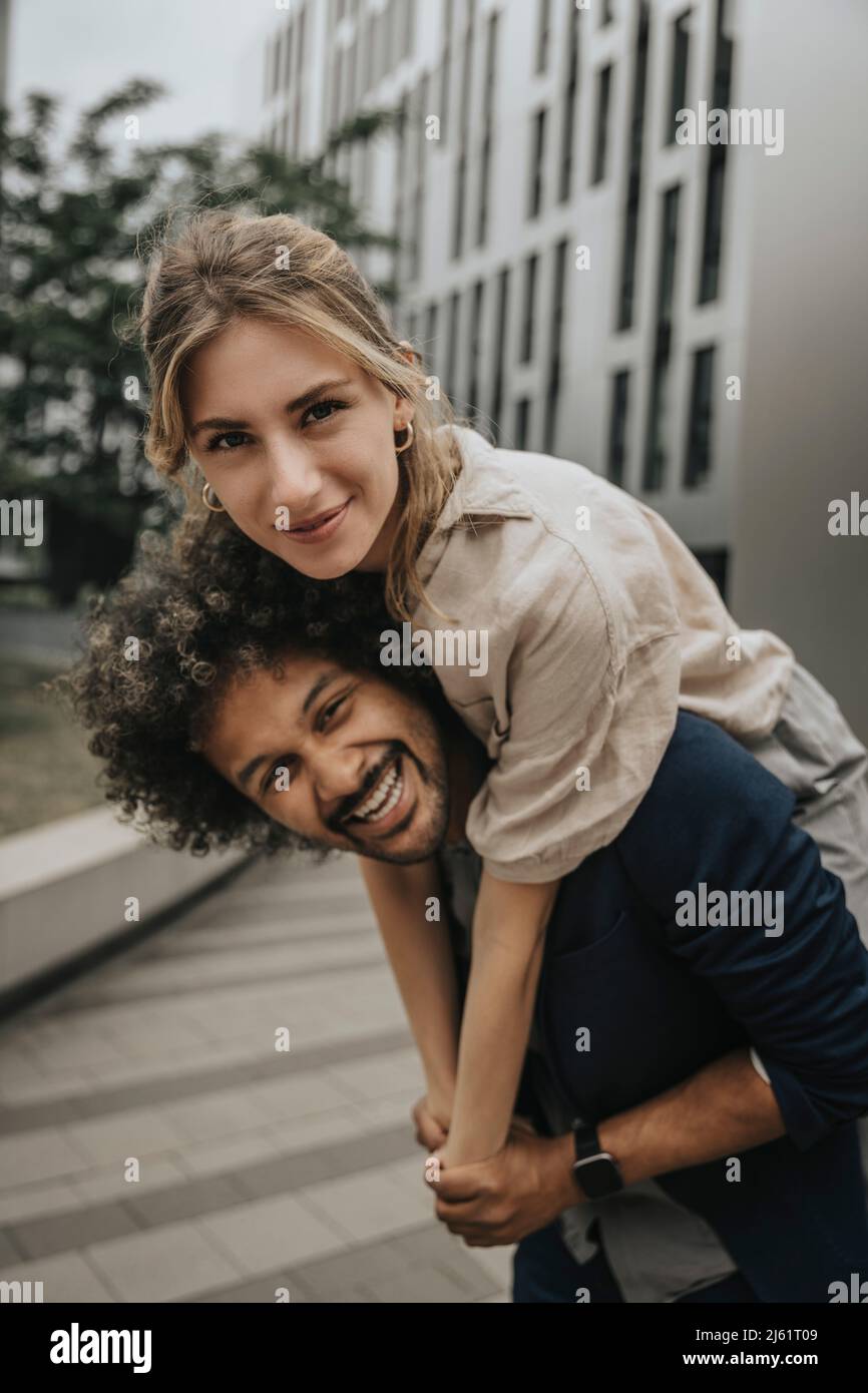 Happy young man giving piggyback ride to girlfriend in front of modern ...
