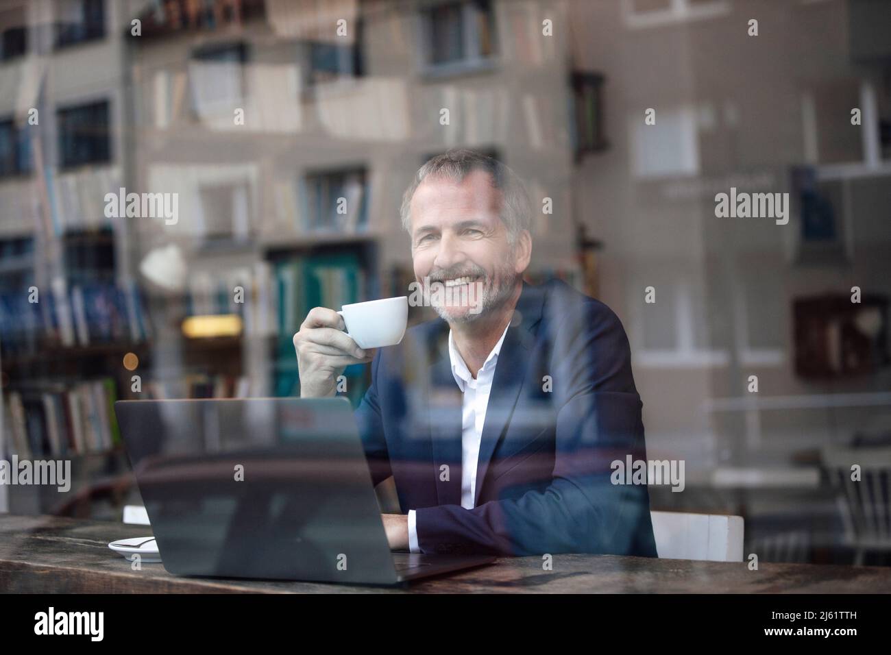 Happy freelancer with coffee cup and laptop seen through glass window ...