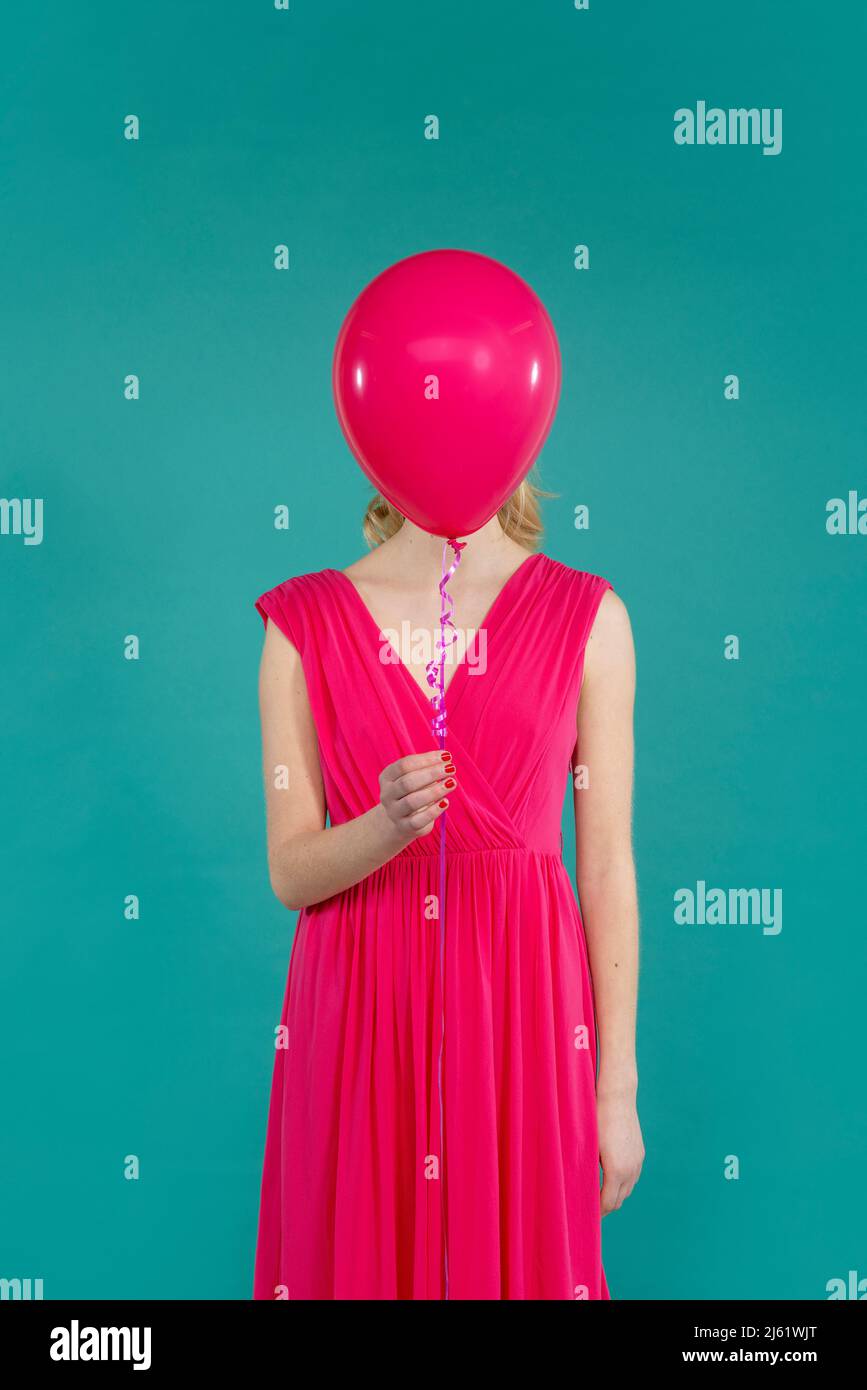 Young woman holding balloon in front of face standing against green ...