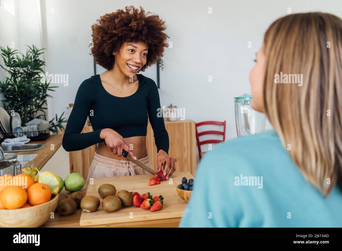 Happy young woman cutting strawberries talking with friend in kitchen ...