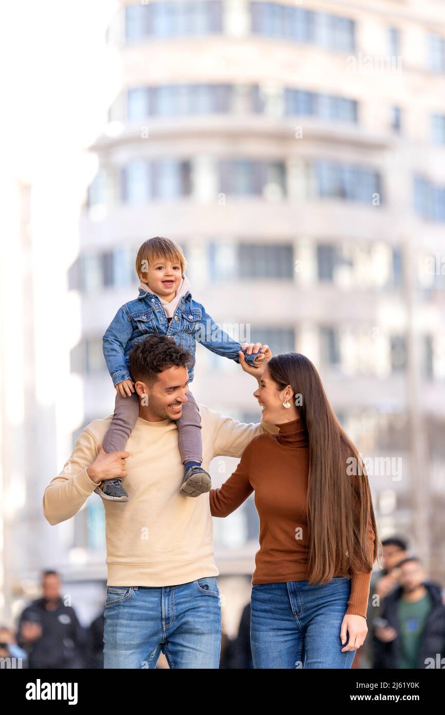Happy man carrying son on shoulder walking with young woman in front of ...