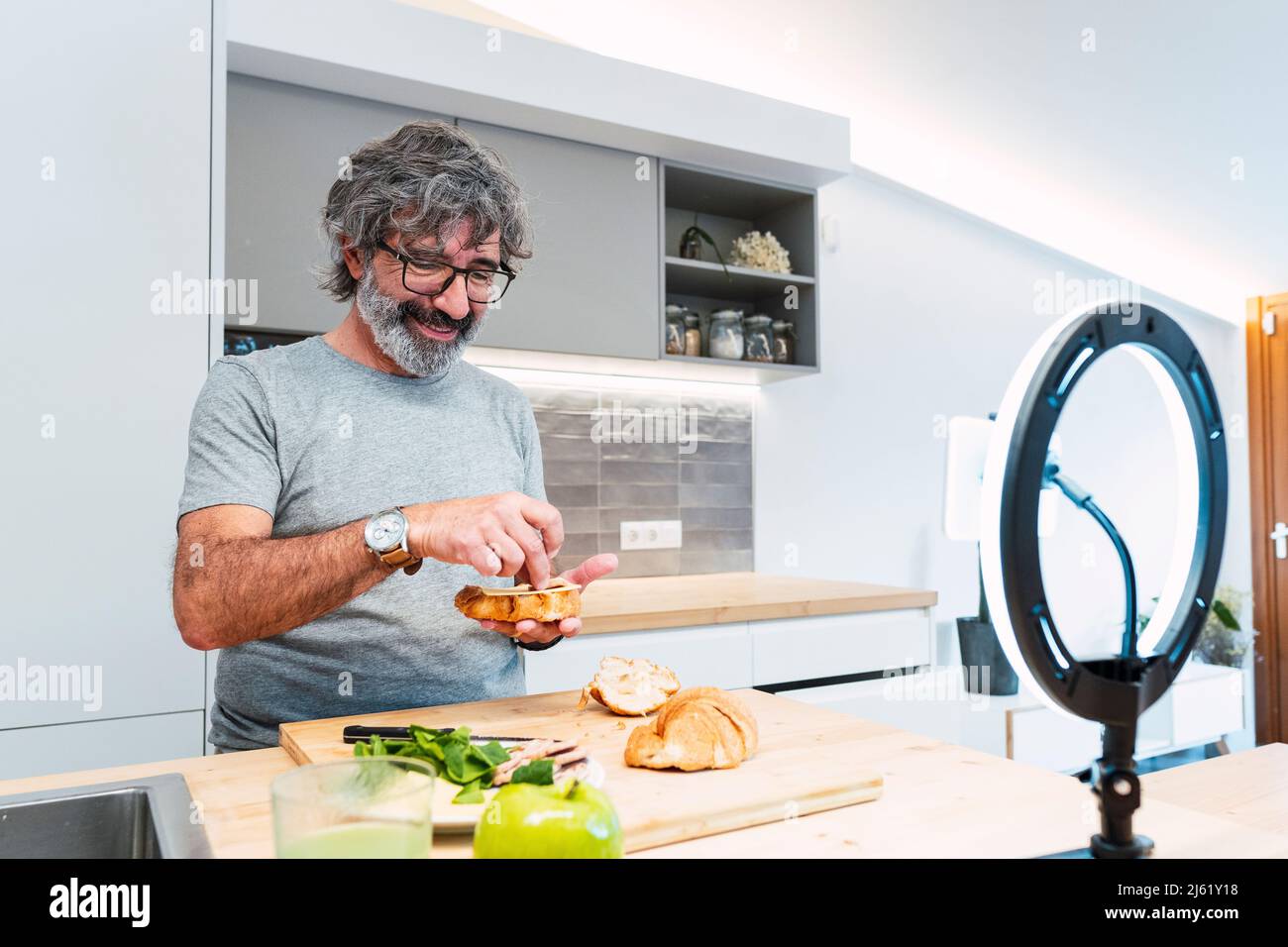Smiling senior man making sandwich with croissant and vlogging through ...
