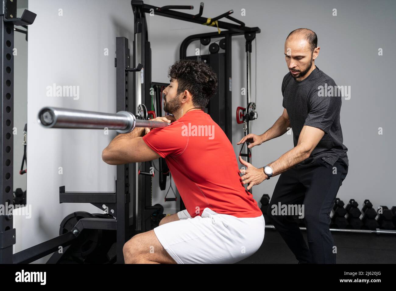 Fitness instructor giving support to young man lifting barbell at ...