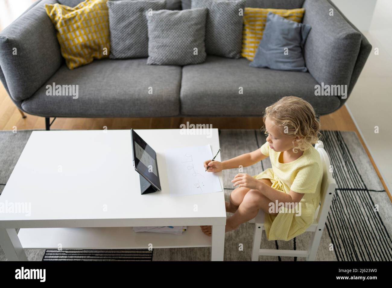 Girl drawing on paper with pencil sitting on chair at table in living ...