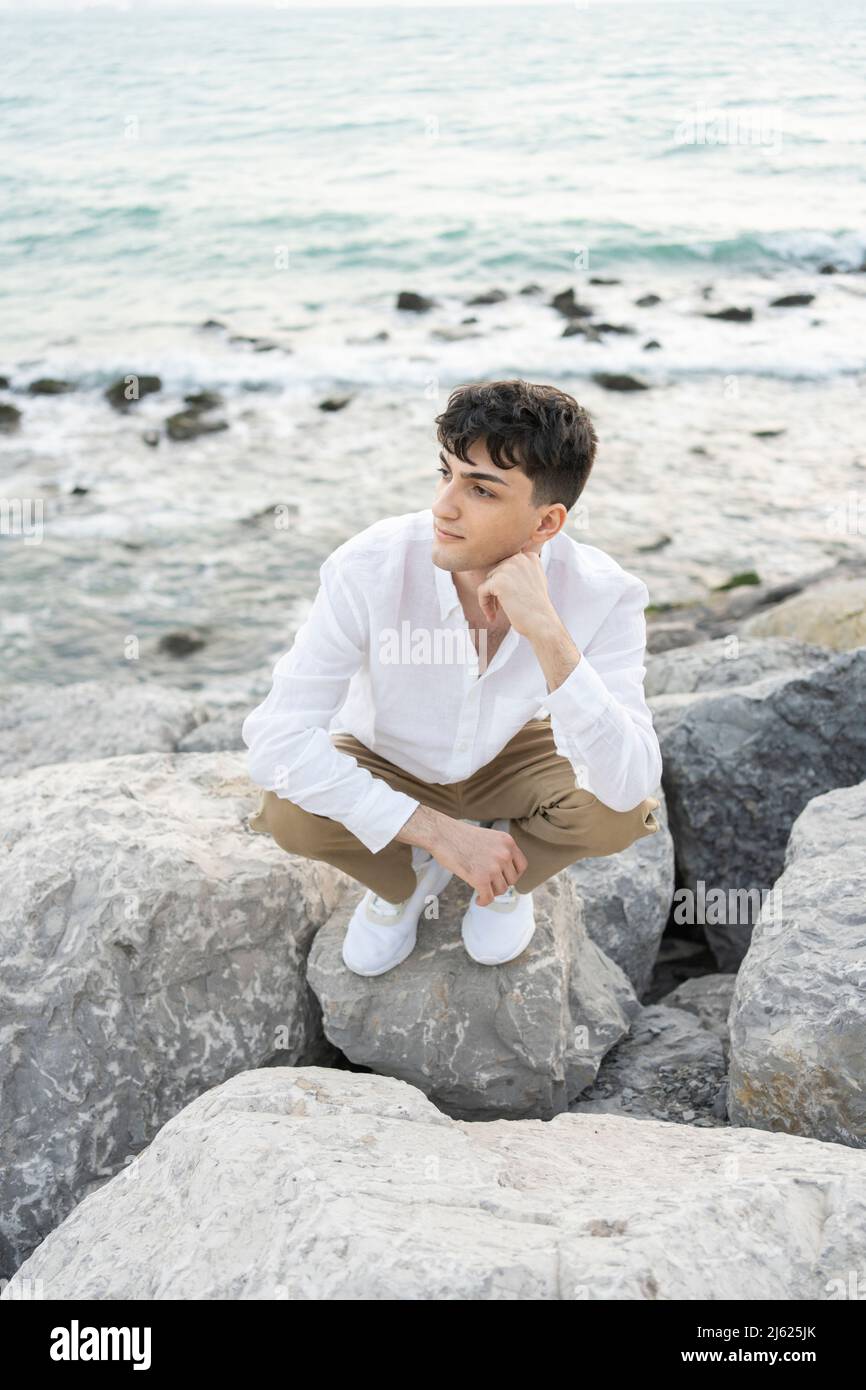 Smiling young man crouching on rock in front on rocky beach Stock Photo ...