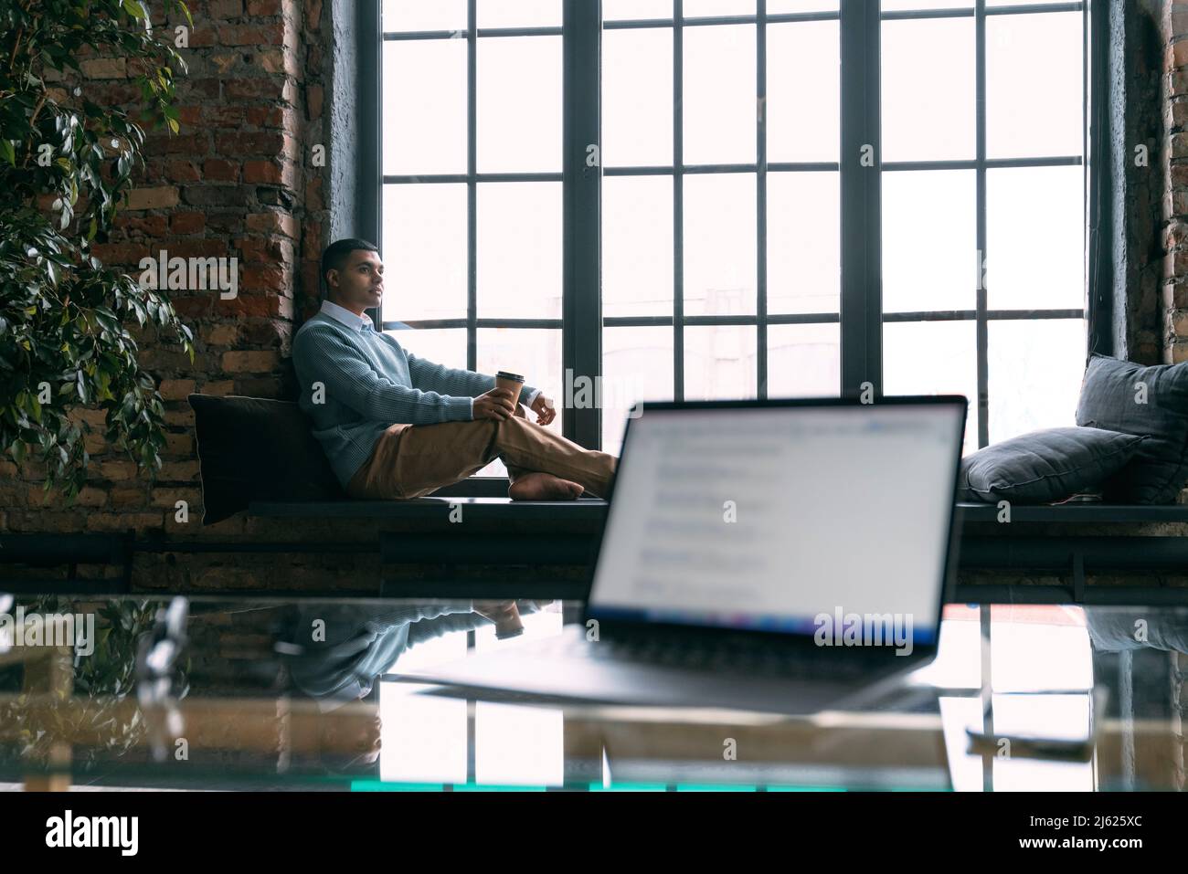 Young freelancer with disposable coffee cup sitting on window sill at ...