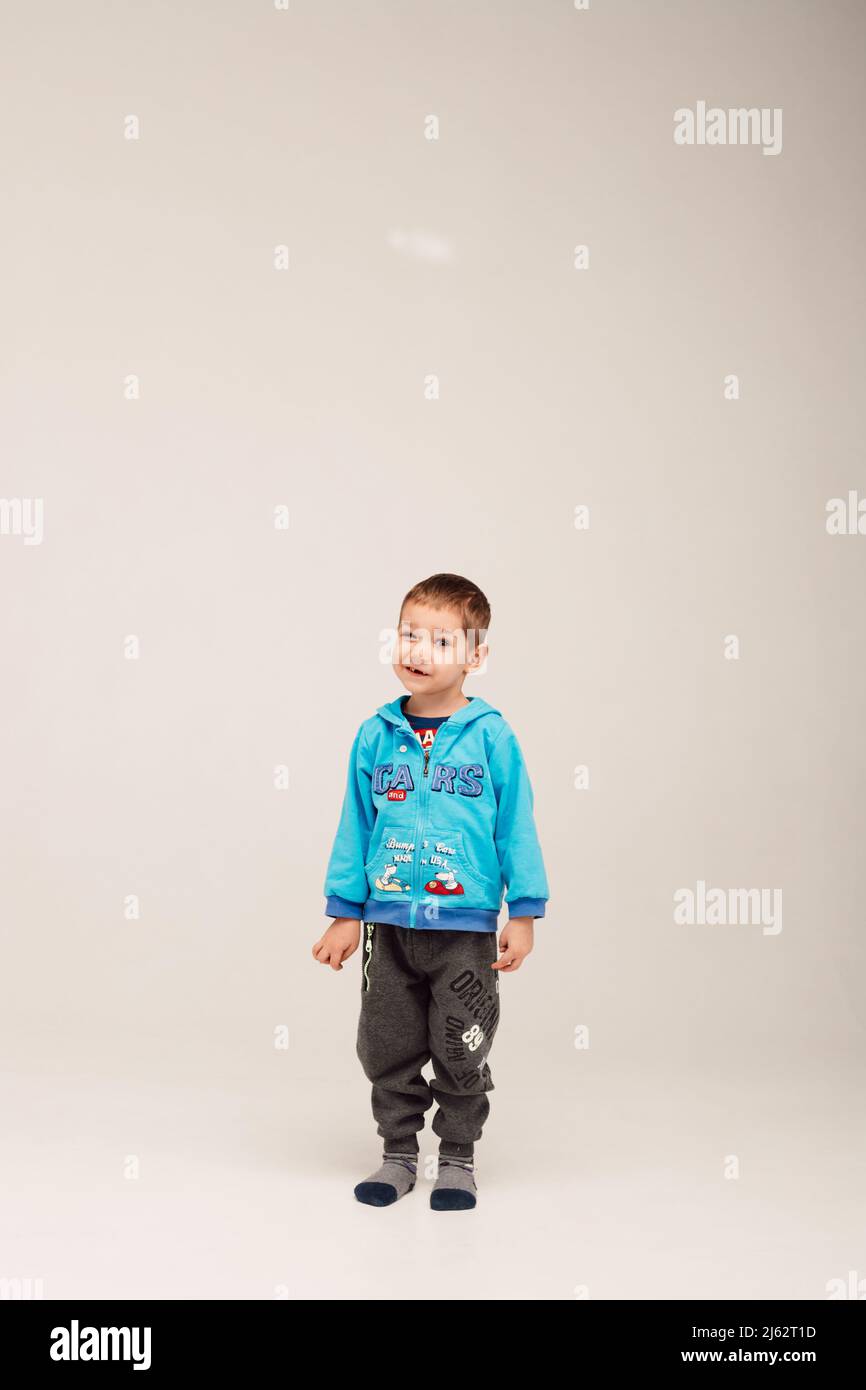 A little boy in a blue jacket stands on a white background and smiles ...