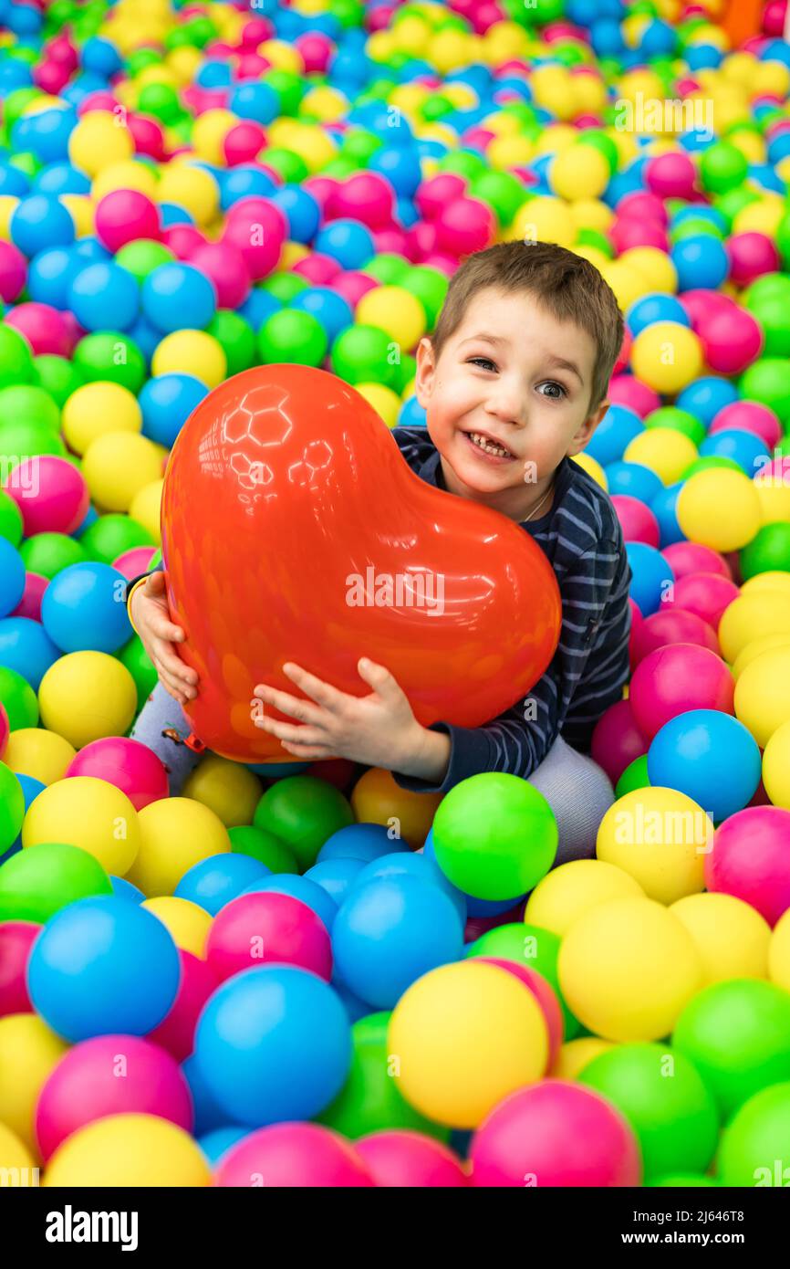A little boy sits in bright balls and holds in his hands a red ball ...