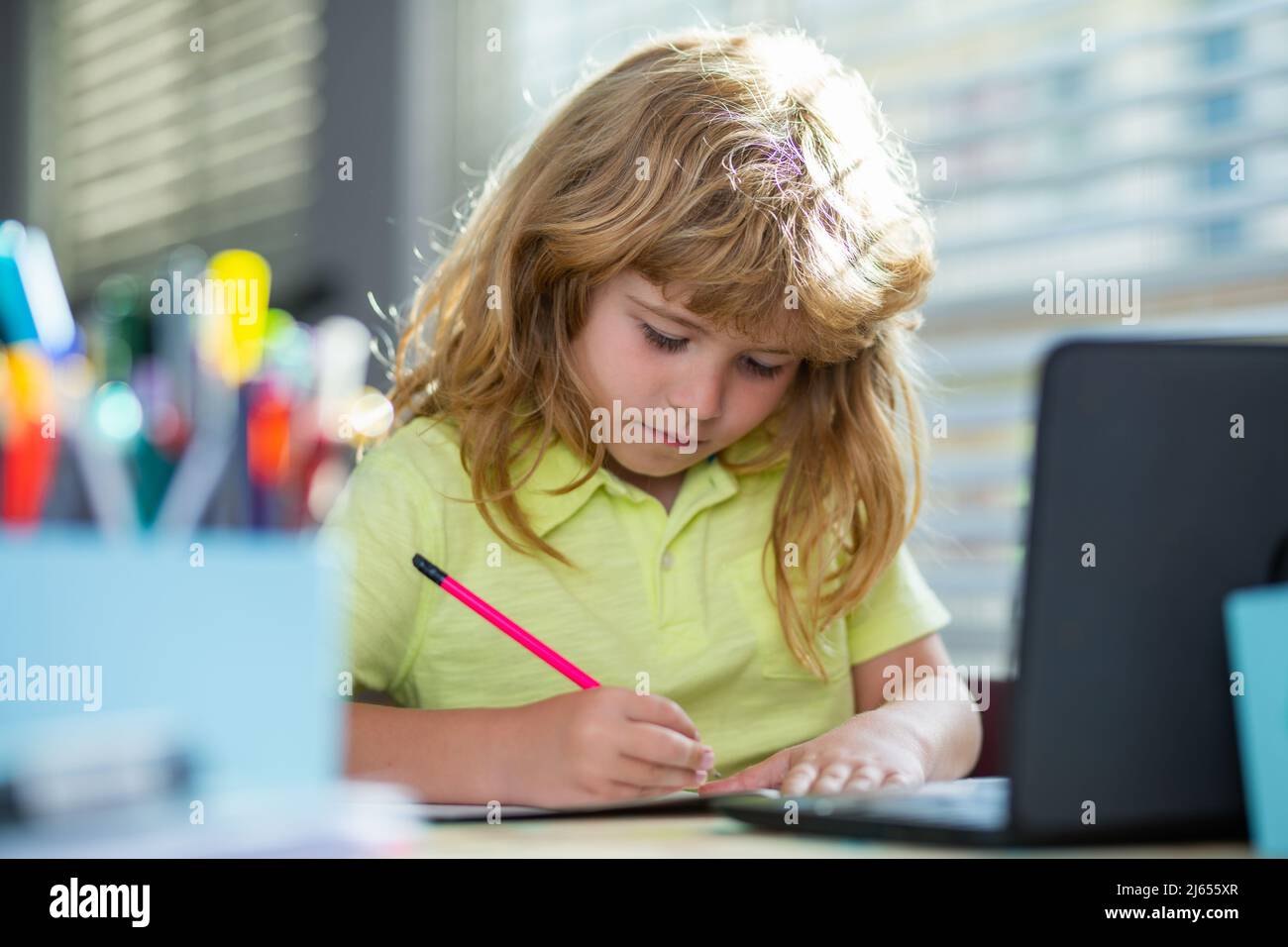 School boy making notes in copybook during online lesson on laptop at ...