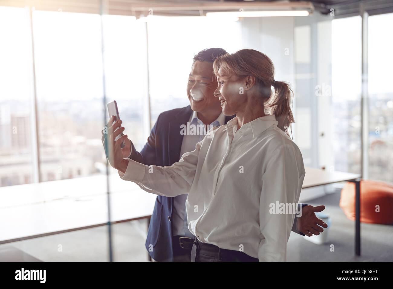 happy colleagues using smartphone for video call while standing near glass wall in modern office ...