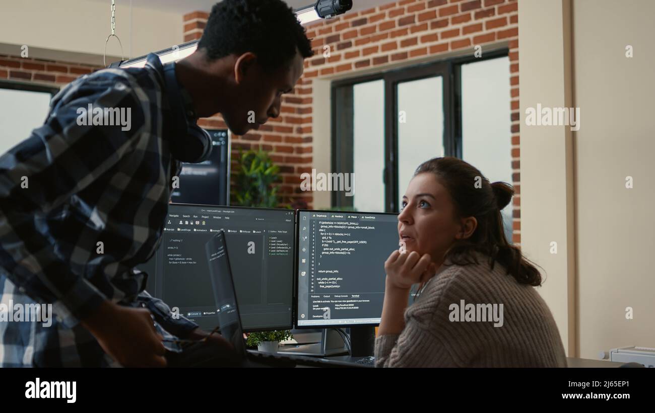 Junior developer sitting down on desk showing laptop with source code to senior dev asking for ...