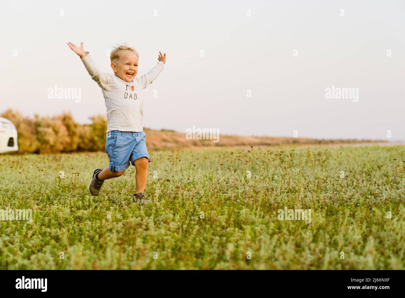 Blonde white boy smiling while running with hands up on summer field ...