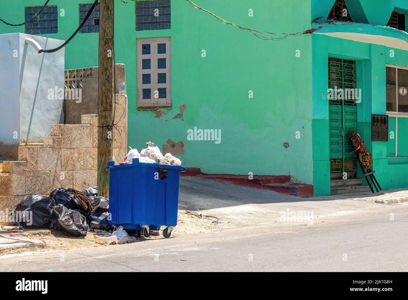 Garbage and garbage bin in a street corner. A wreath of flower is seen ...