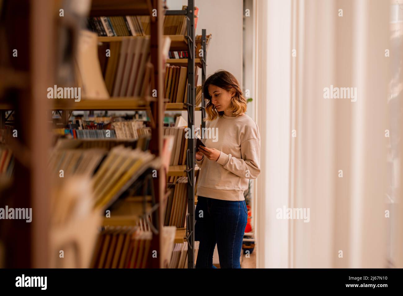 College girl reading a book while standing besides bookshelves Stock ...