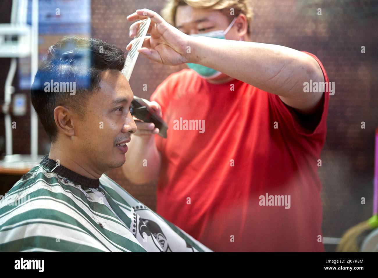 Fat barber with facial mask cutting the hair of an asian client in a ...