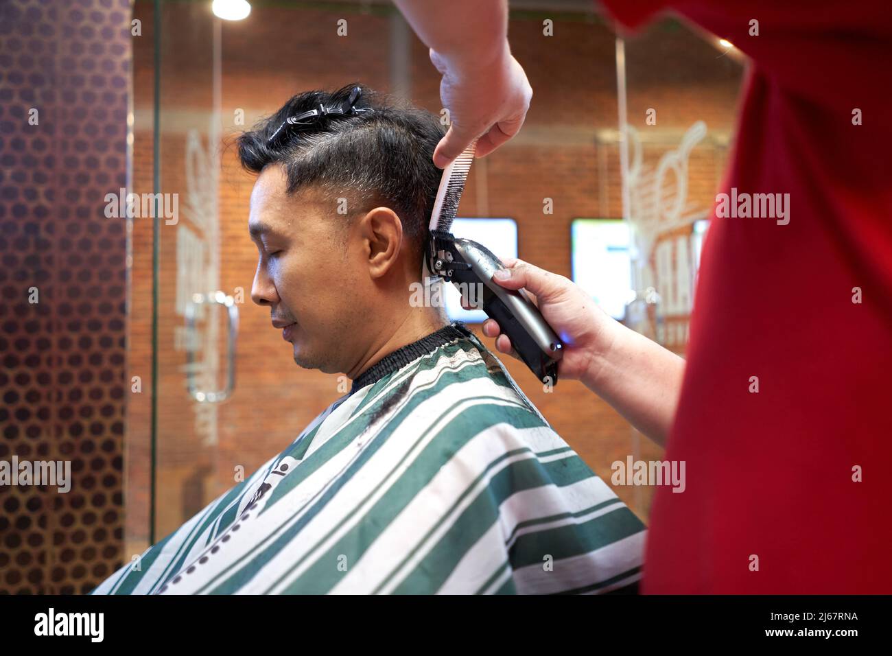 Asian man sits in a barber's shop while his hair is cut with an ...