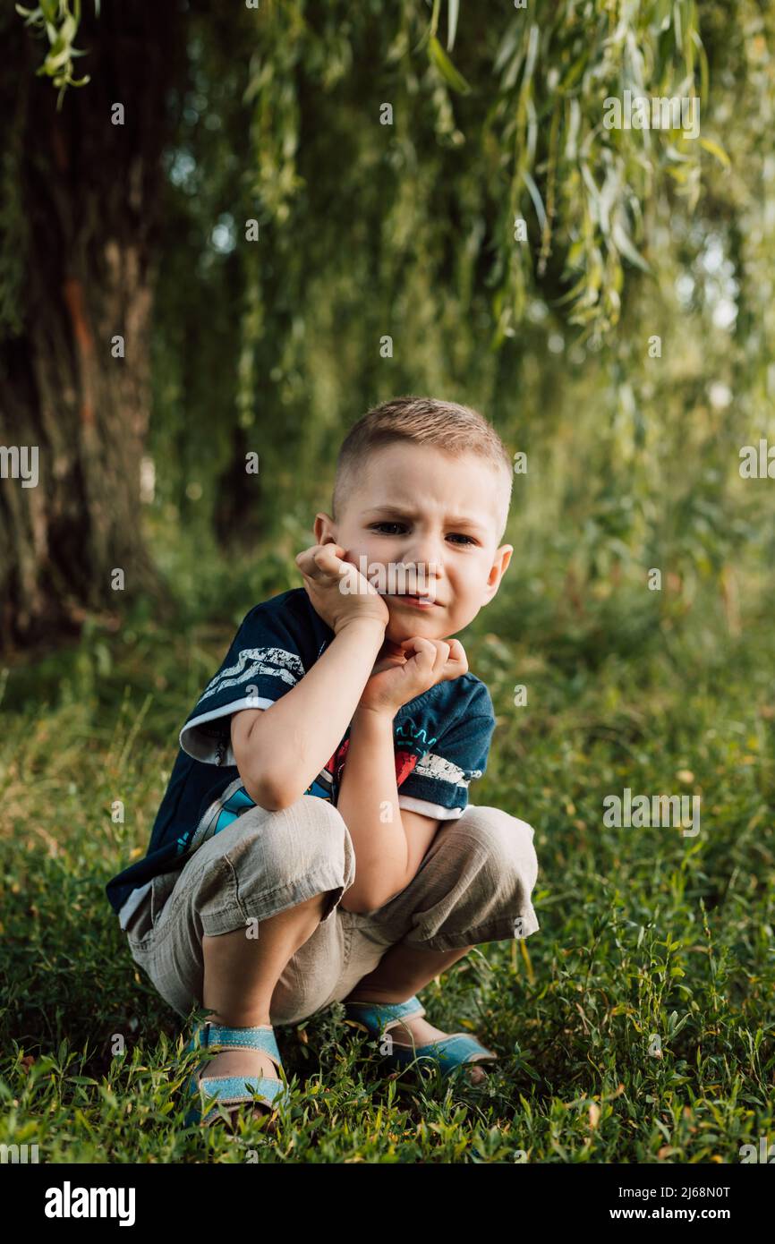 A little boy crouched under a tree, his hand propping up his head and ...
