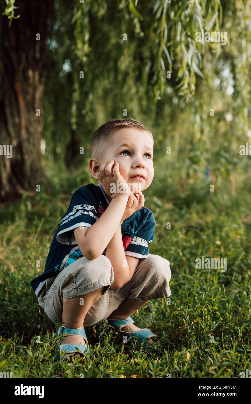 A little boy crouched under a tree, his hand propping up his head and ...