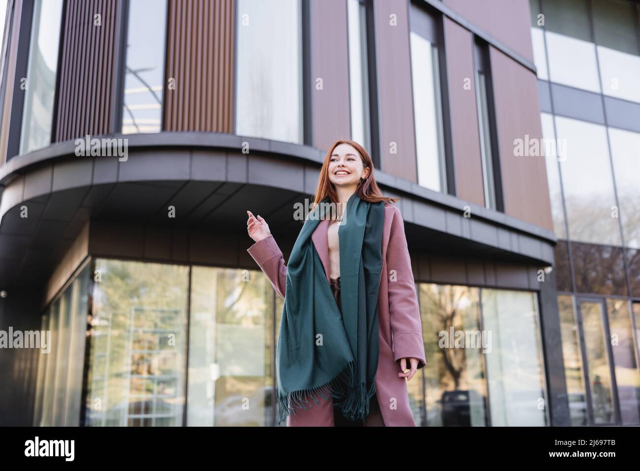 low angle view of joyful redhead woman in coat with scarf near modern ...