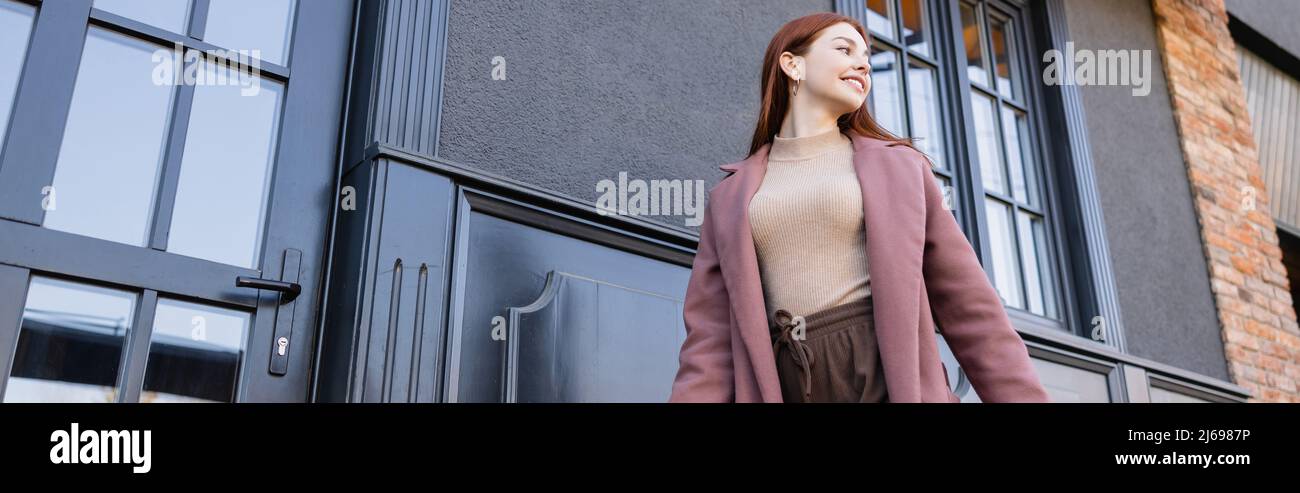 low angle view of happy redhead woman in stylish coat walking near ...