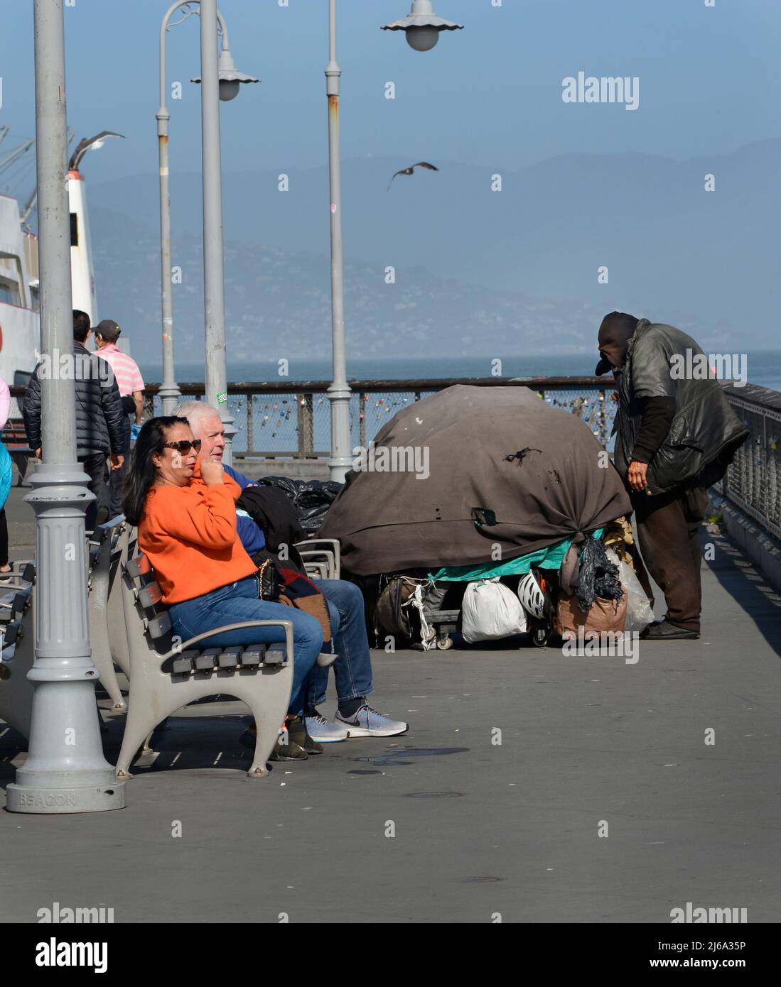 Tourists sit on a pier near the makeshift shelter of a homeless man in ...