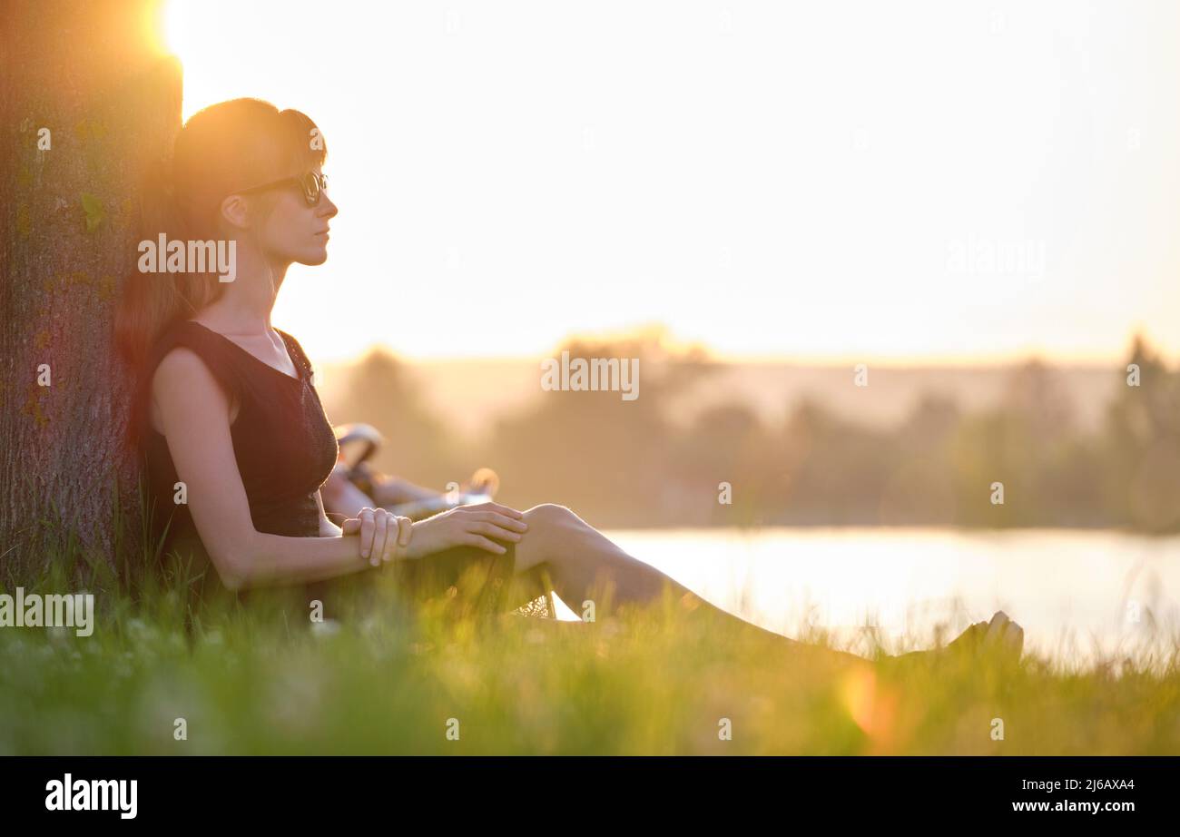 Lonely woman sitting alone on green grass lawn leaning to tree trunk on ...