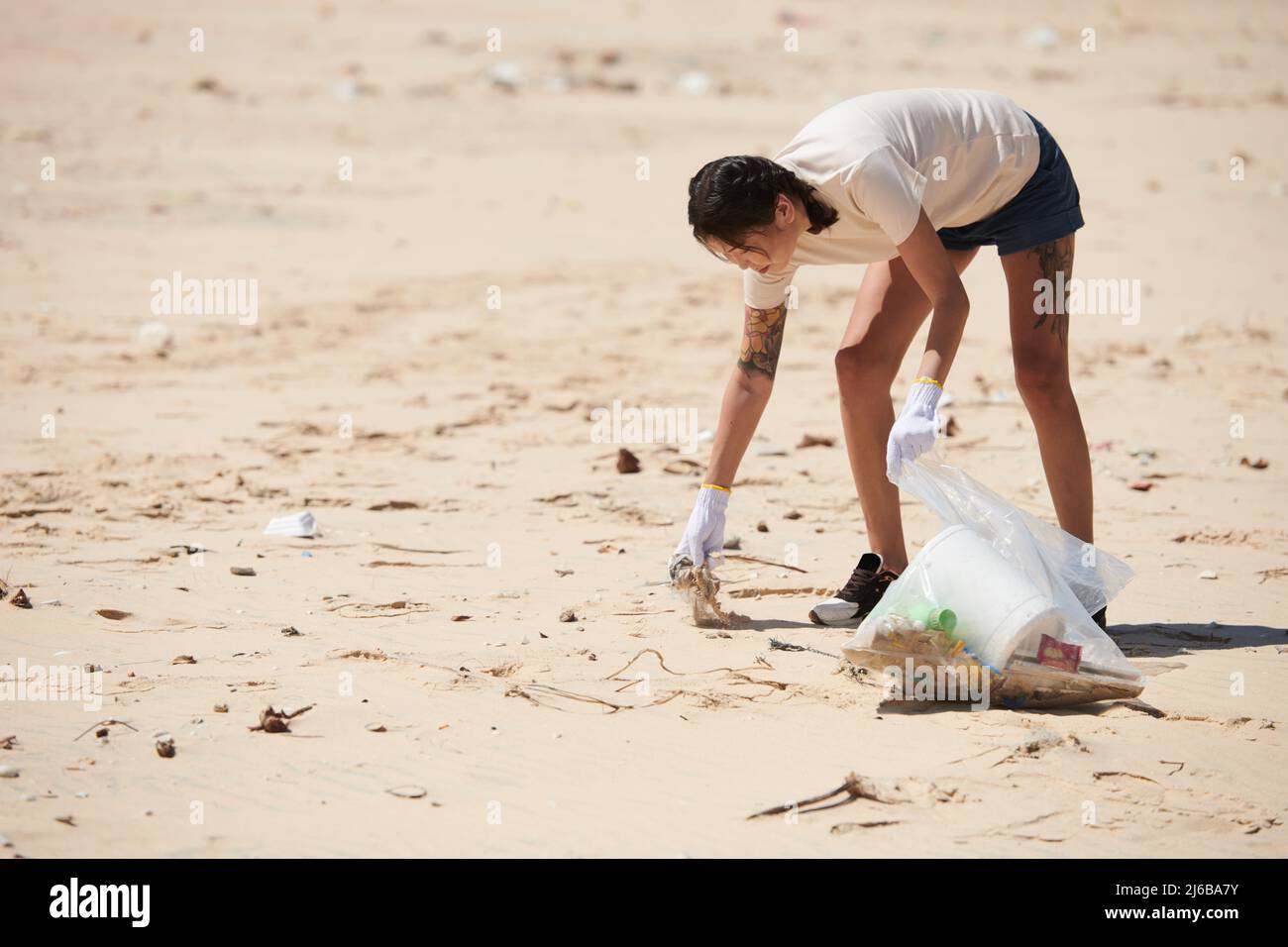 Young woman wearing textile gloves when picking up trash on sandy beach ...