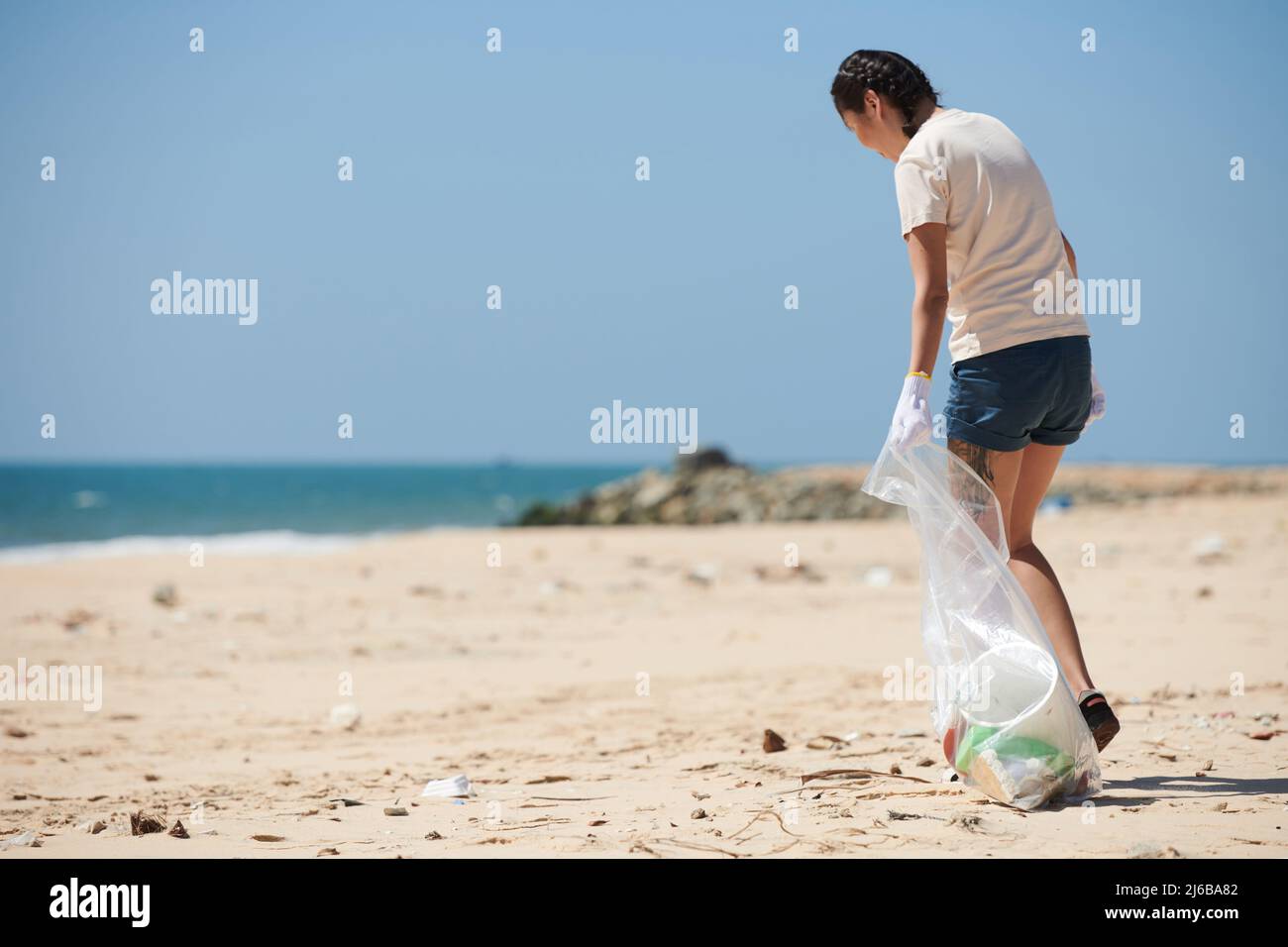Young woman walking on beach with big plastic bag and collecting trash ...