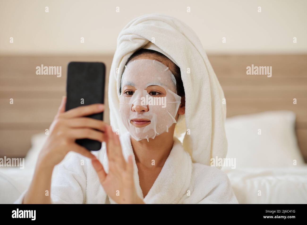 Young woman with sheet mask on face texting friends or checking social ...