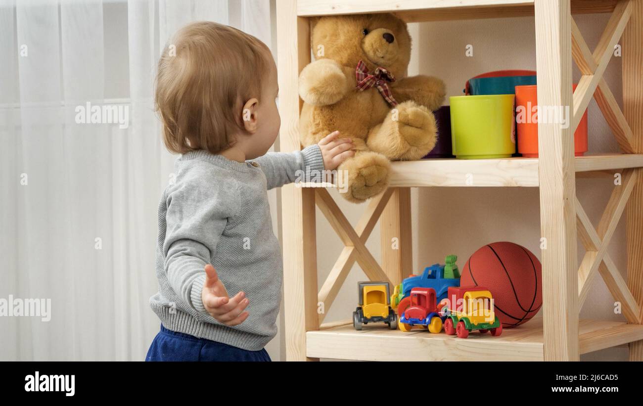 Cute baby boy taking teddy bear toy from bookshelf in playroom. Child ...