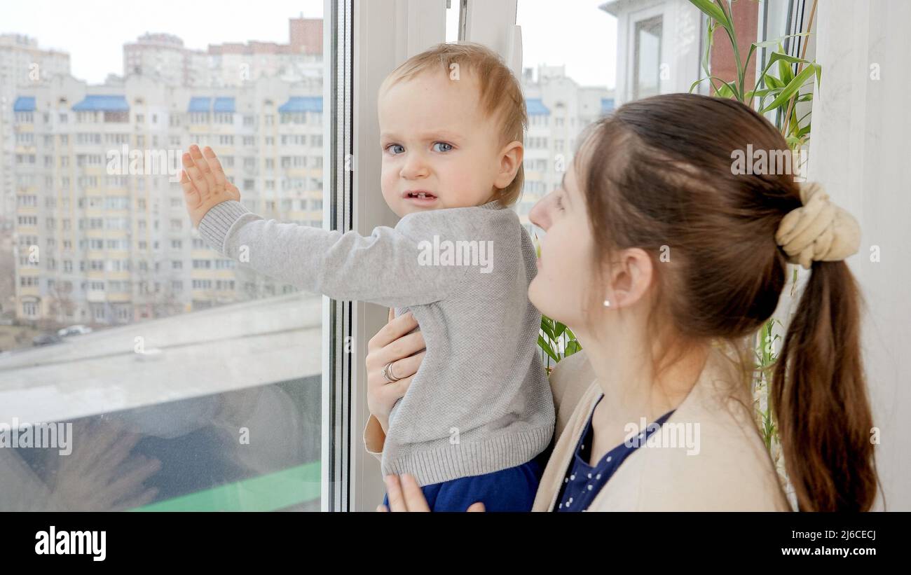 Young mother holding her baby son trying to open window on high floor ...
