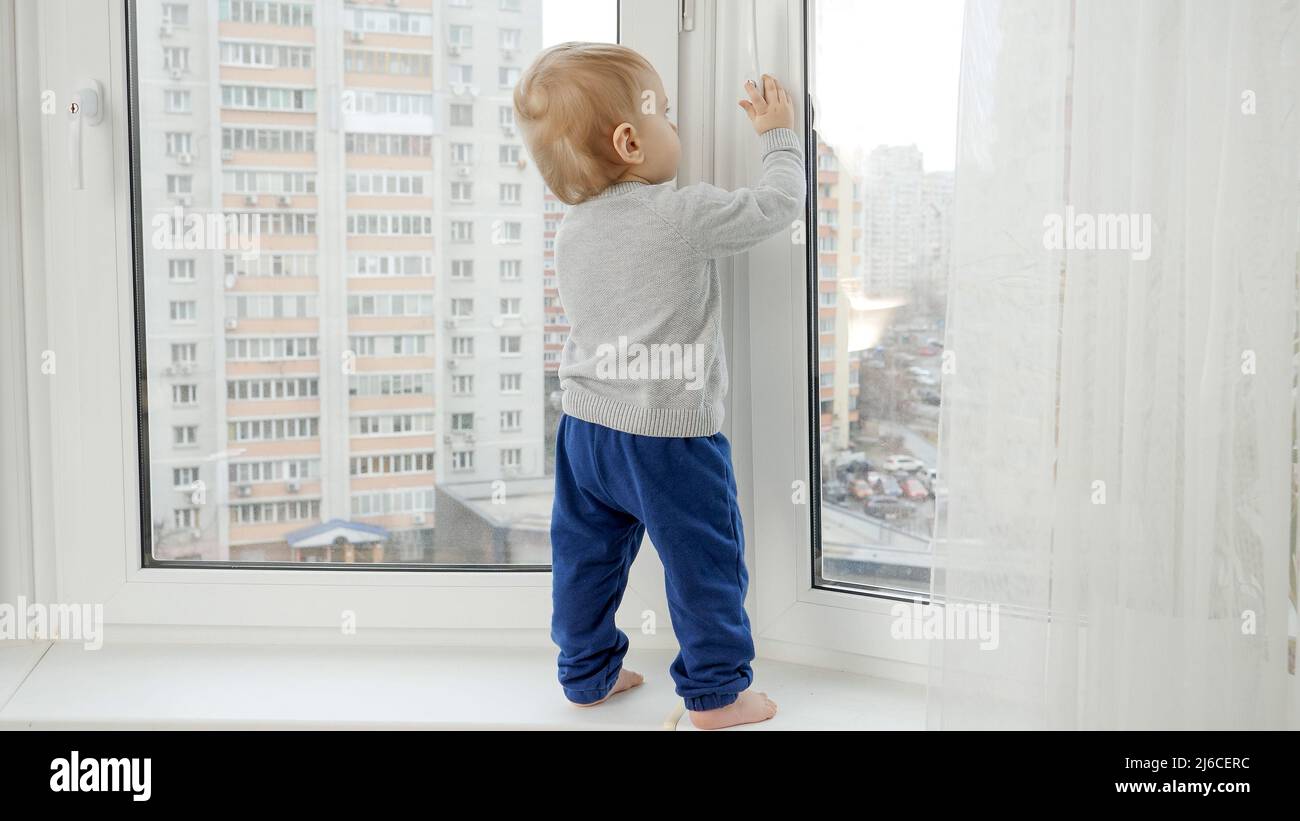 Little baby boy standing on windowsill and pulling window handle. Baby ...