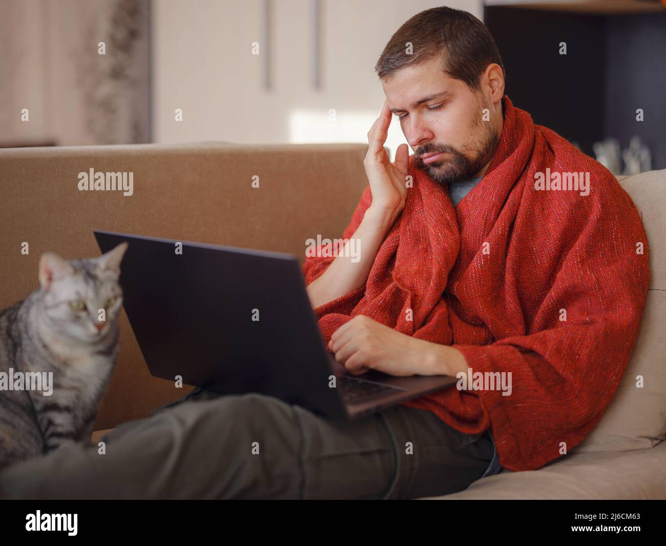 Ill man covered in warm blanket looking at laptop screen, sitting on ...