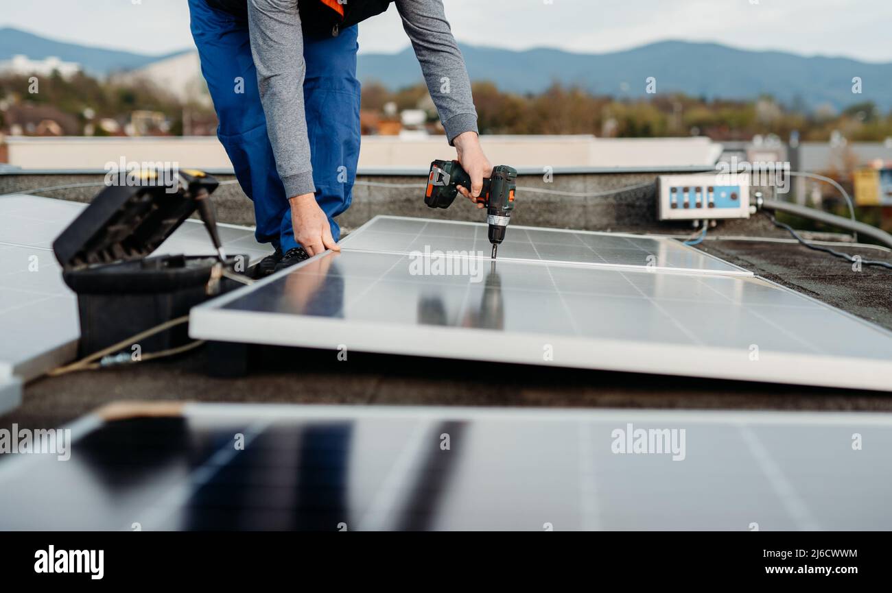 Solar panel technician with drill installing solar panels on roof on family house a sunny day ...