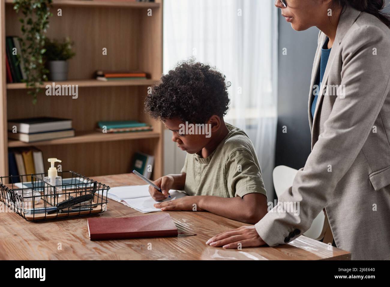 Tutor in jacket standing at table and watching how black boy doing task ...