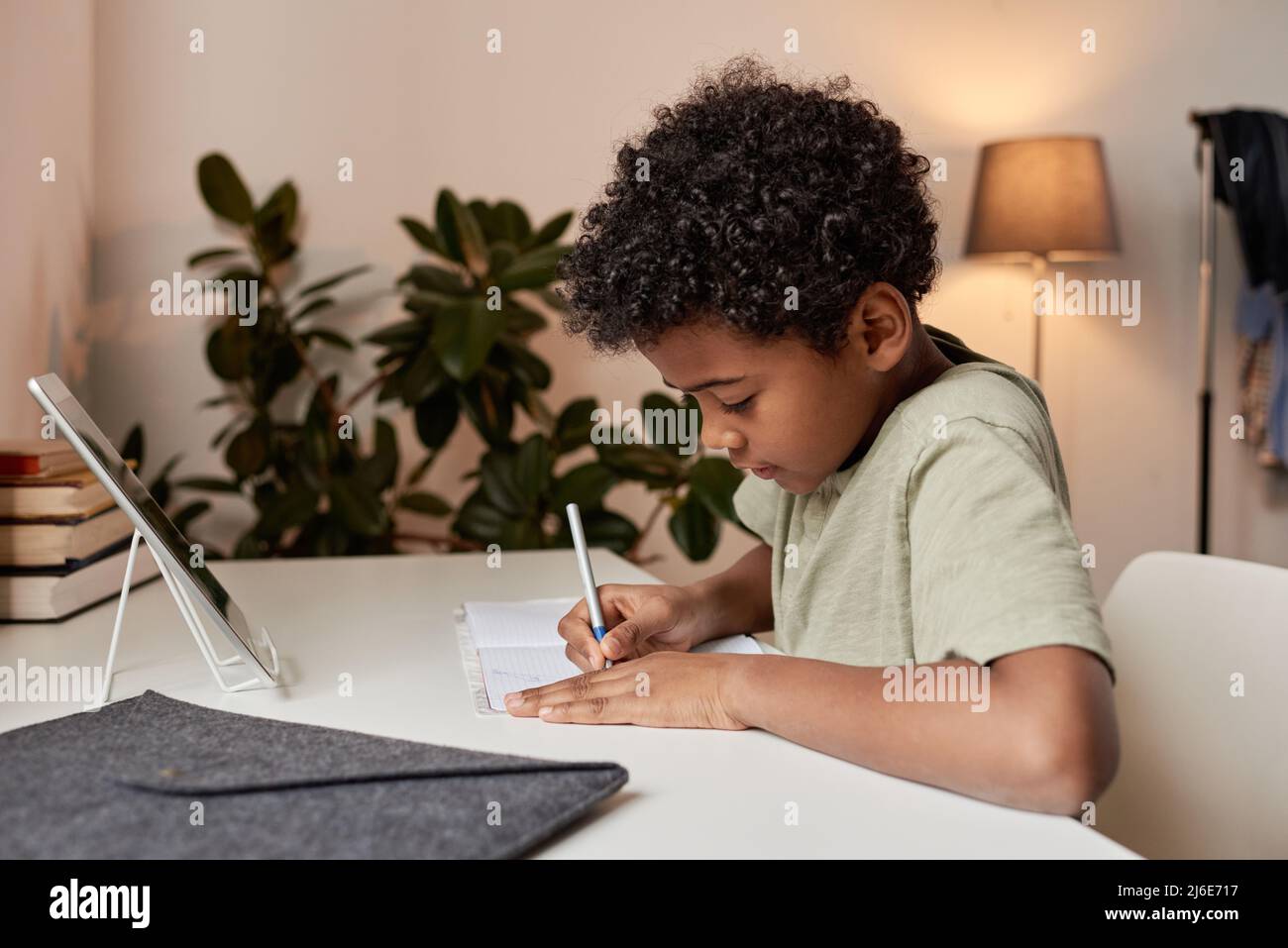 Serious curly-haired black boy sitting at desk and making notes in ...
