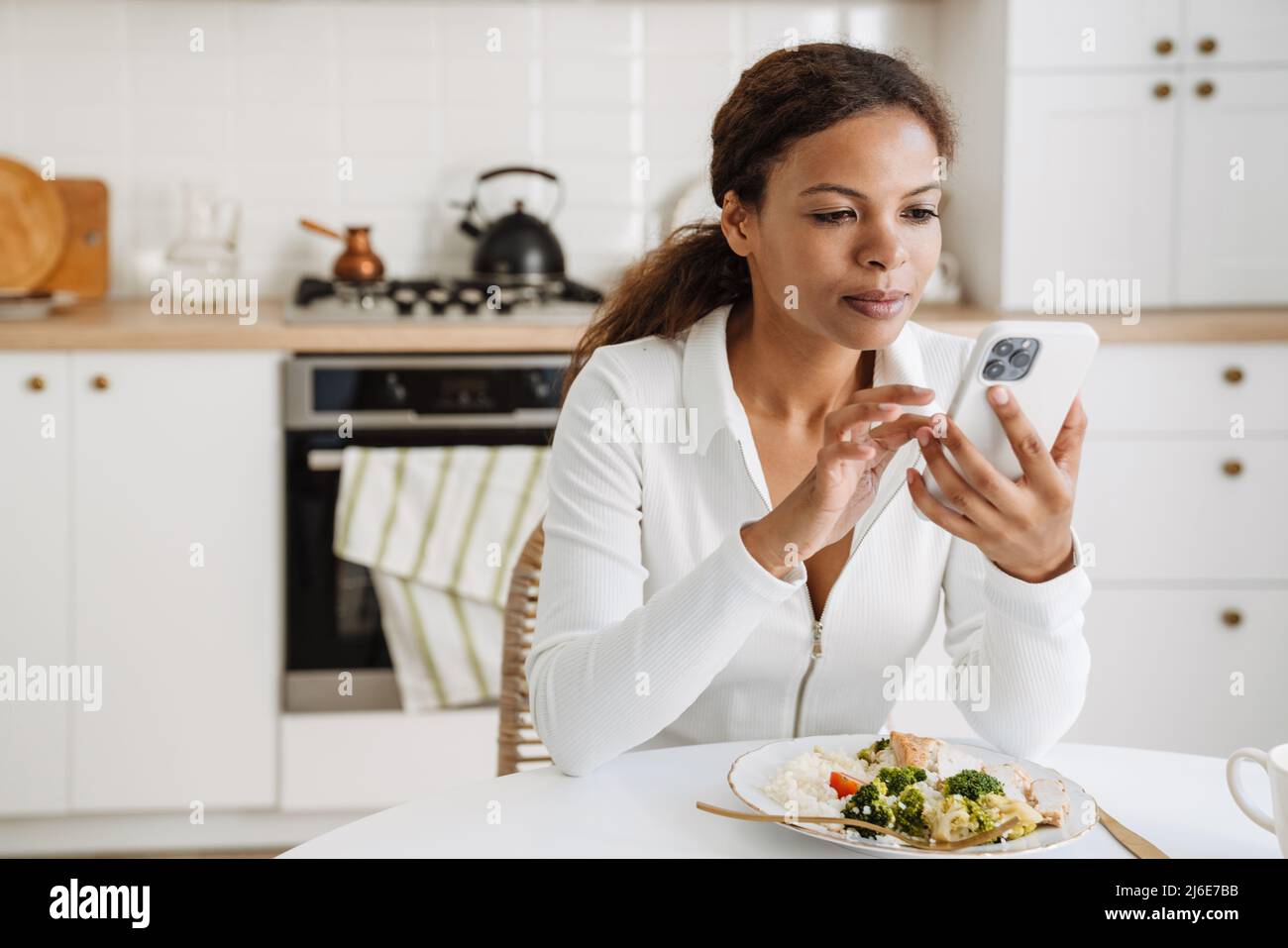 Young black woman using cellphone while having lunch in kitchen at home ...