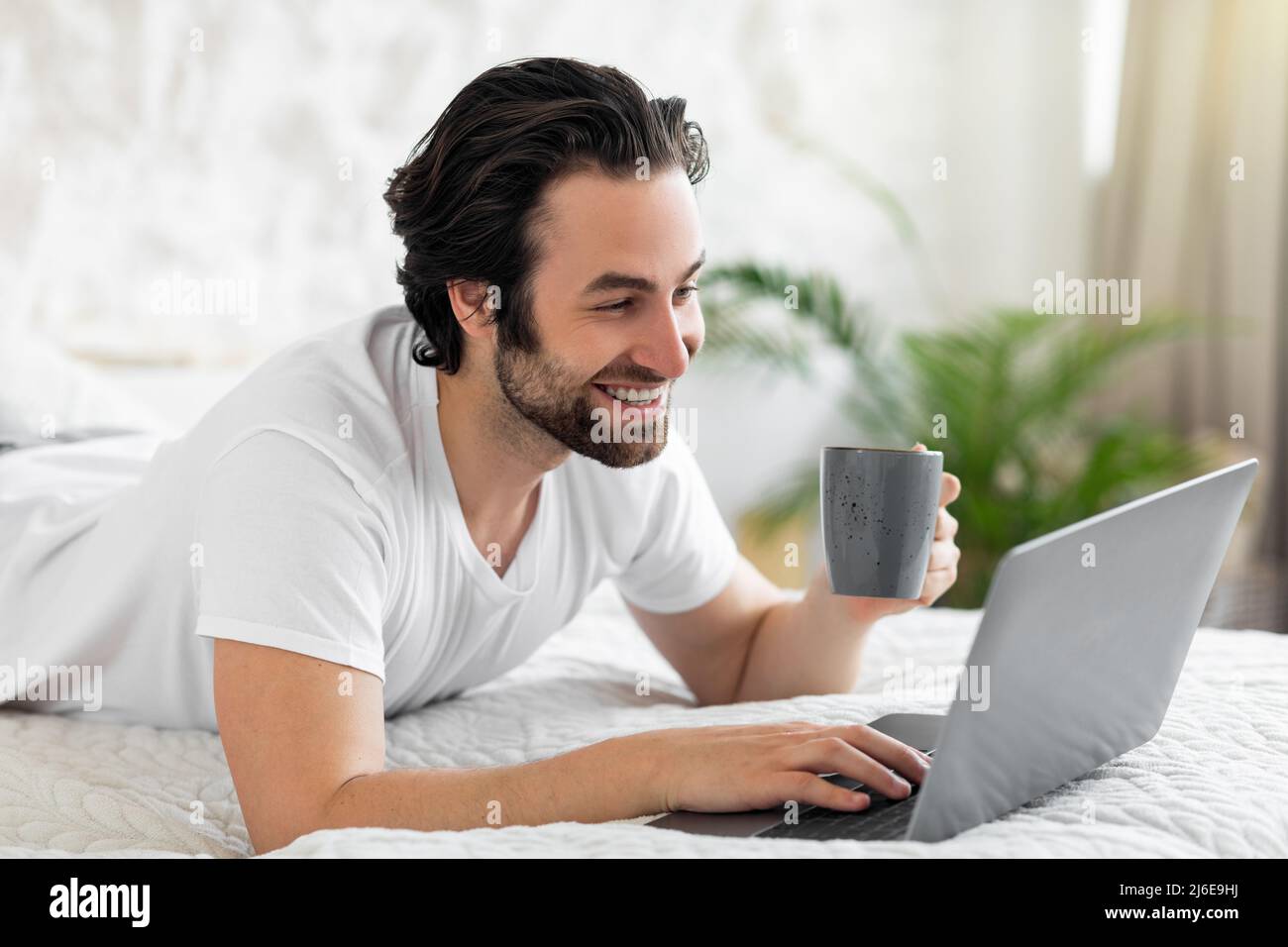 Young millennial guy using laptop in bed, drinking coffee Stock Photo ...