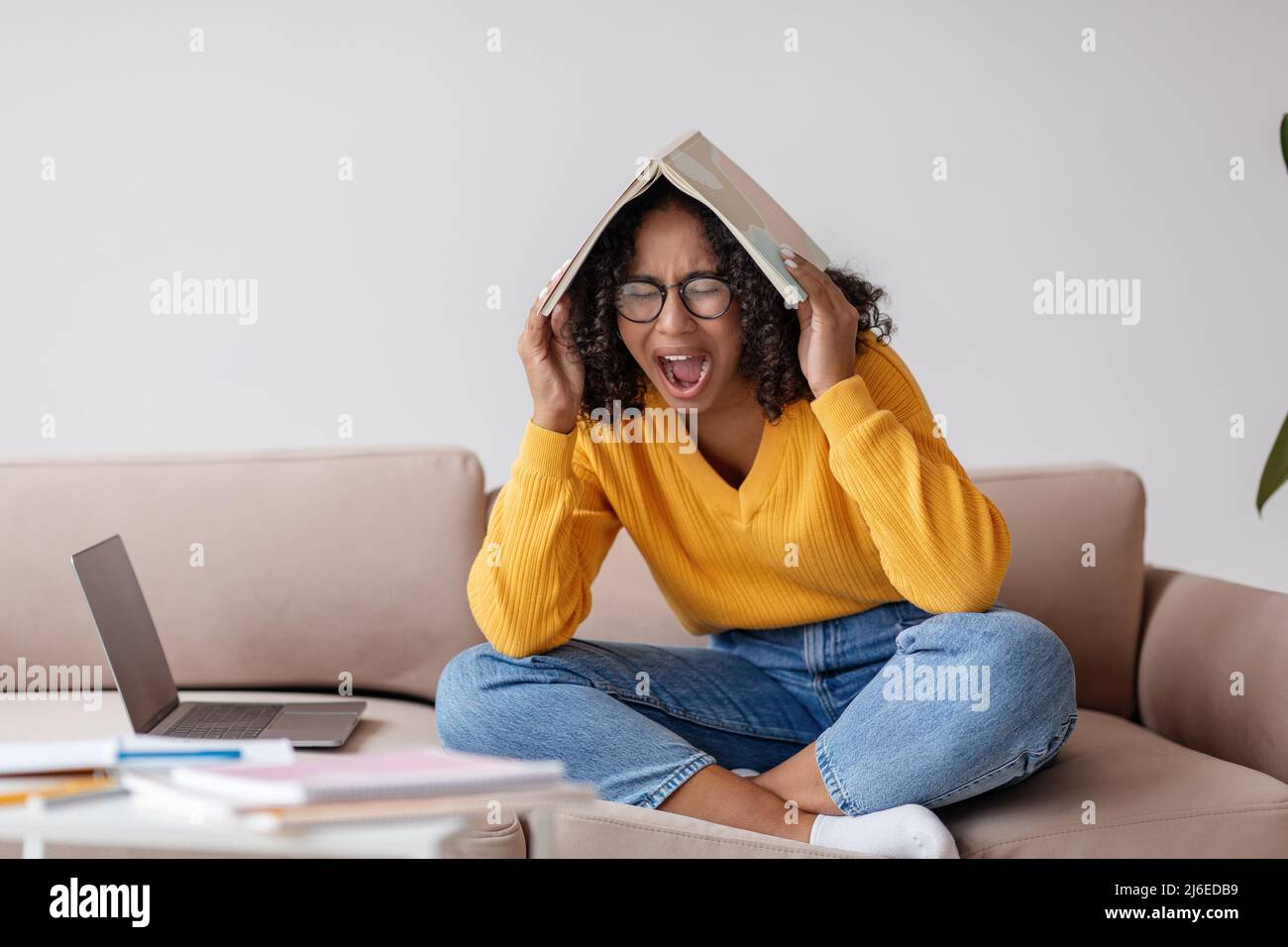 Young black woman shouting with book on her head near laptop computer ...