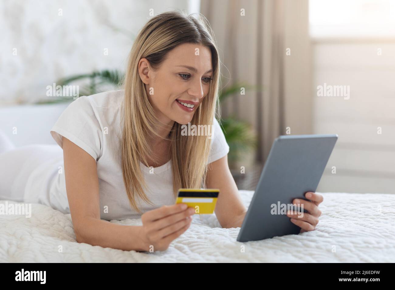 Positive woman laying in bed with tablet and credit card Stock Photo ...