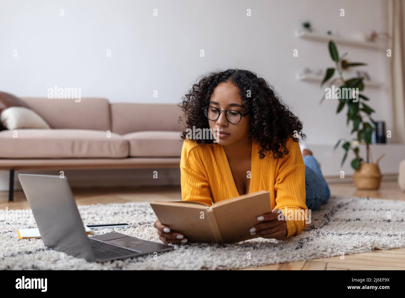 Focused young black lady reading book, lying on floor near laptop ...