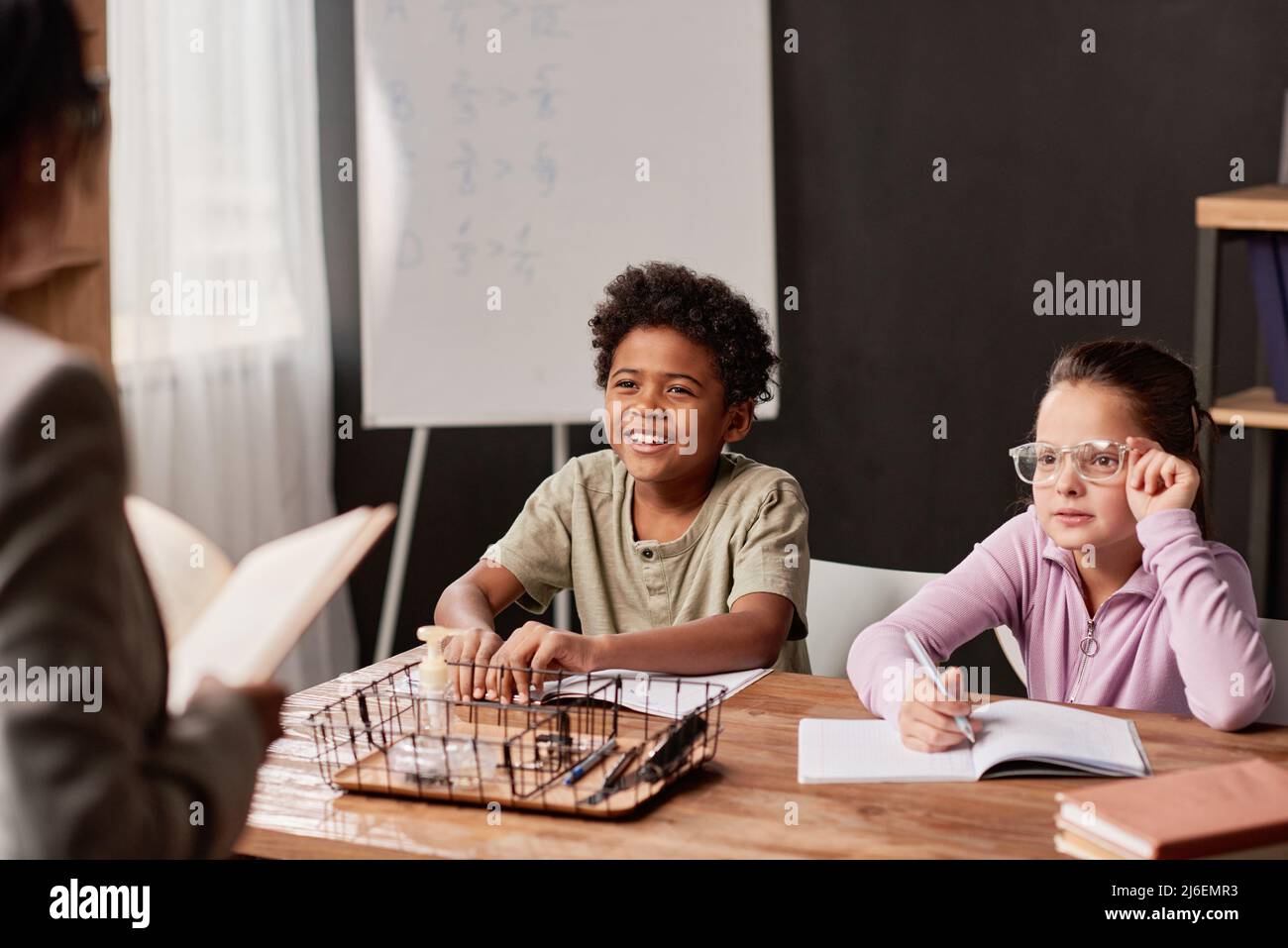 Smiling black boy and serious nerd girl sitting at table with workbook while listening to ...